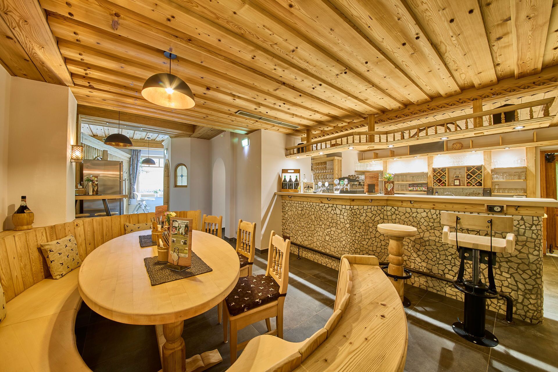 Um interior de madeira acolhedor, com uma mesa de jantar redonda, cadeiras de madeira, uma parede de destaque em pedra e um espaço de bar com prateleiras cheias de copos e garrafas.