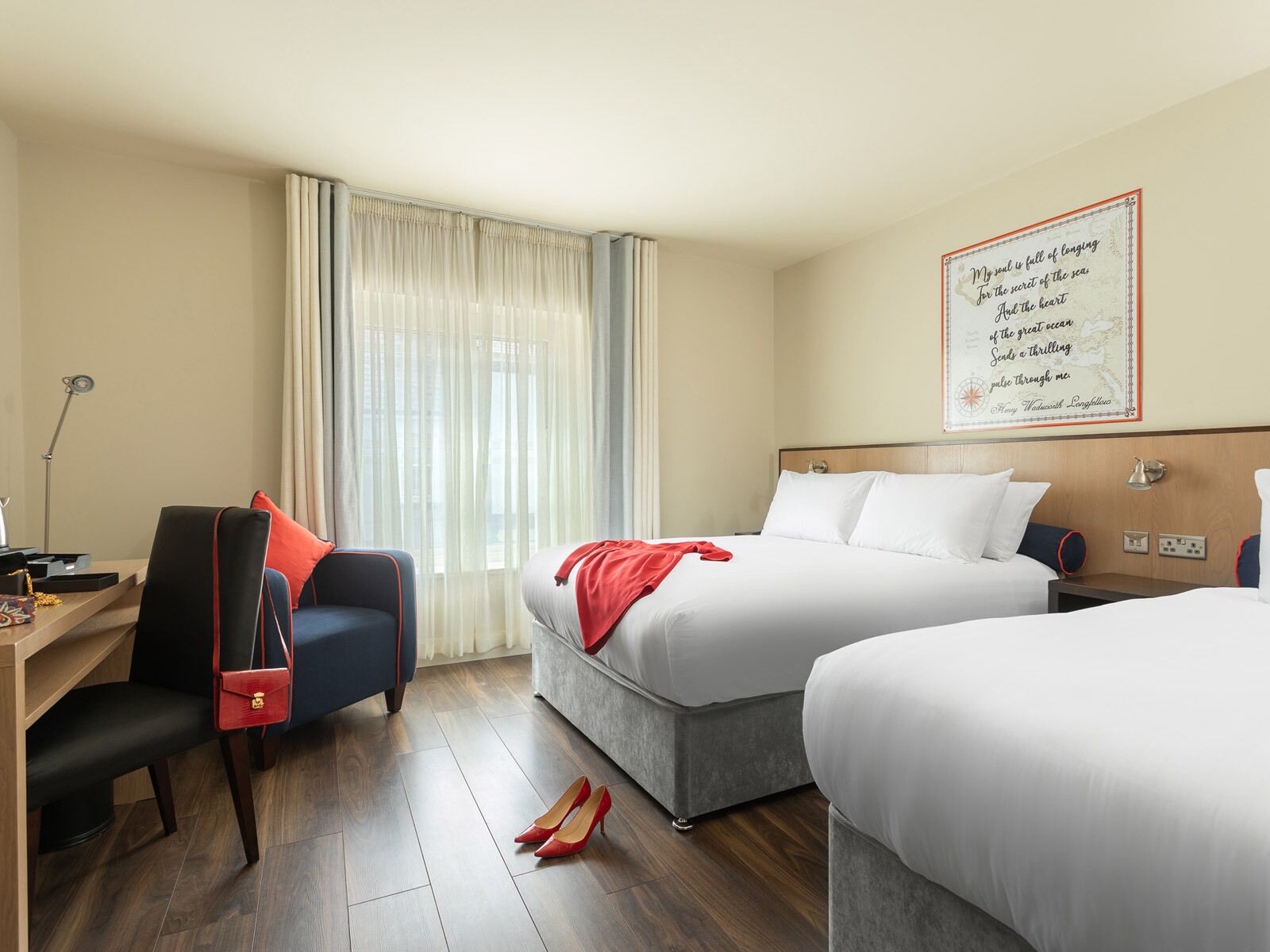 Quarto de hotel com duas camas, uma escrivaninha, uma cadeira e uma janela com cortinas.