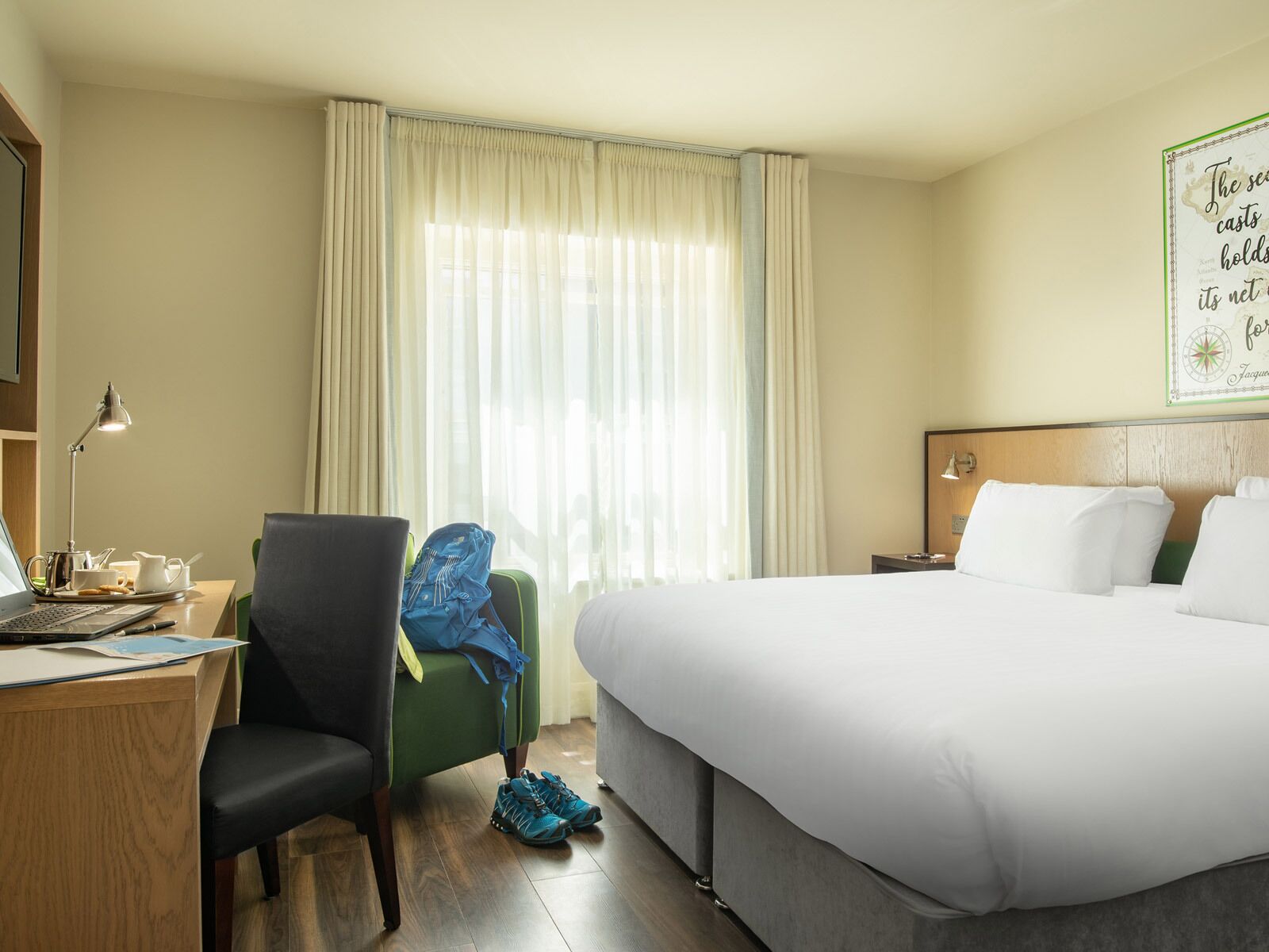 Quarto de hotel com cama, escrivaninha, cadeira e janela com cortinas.