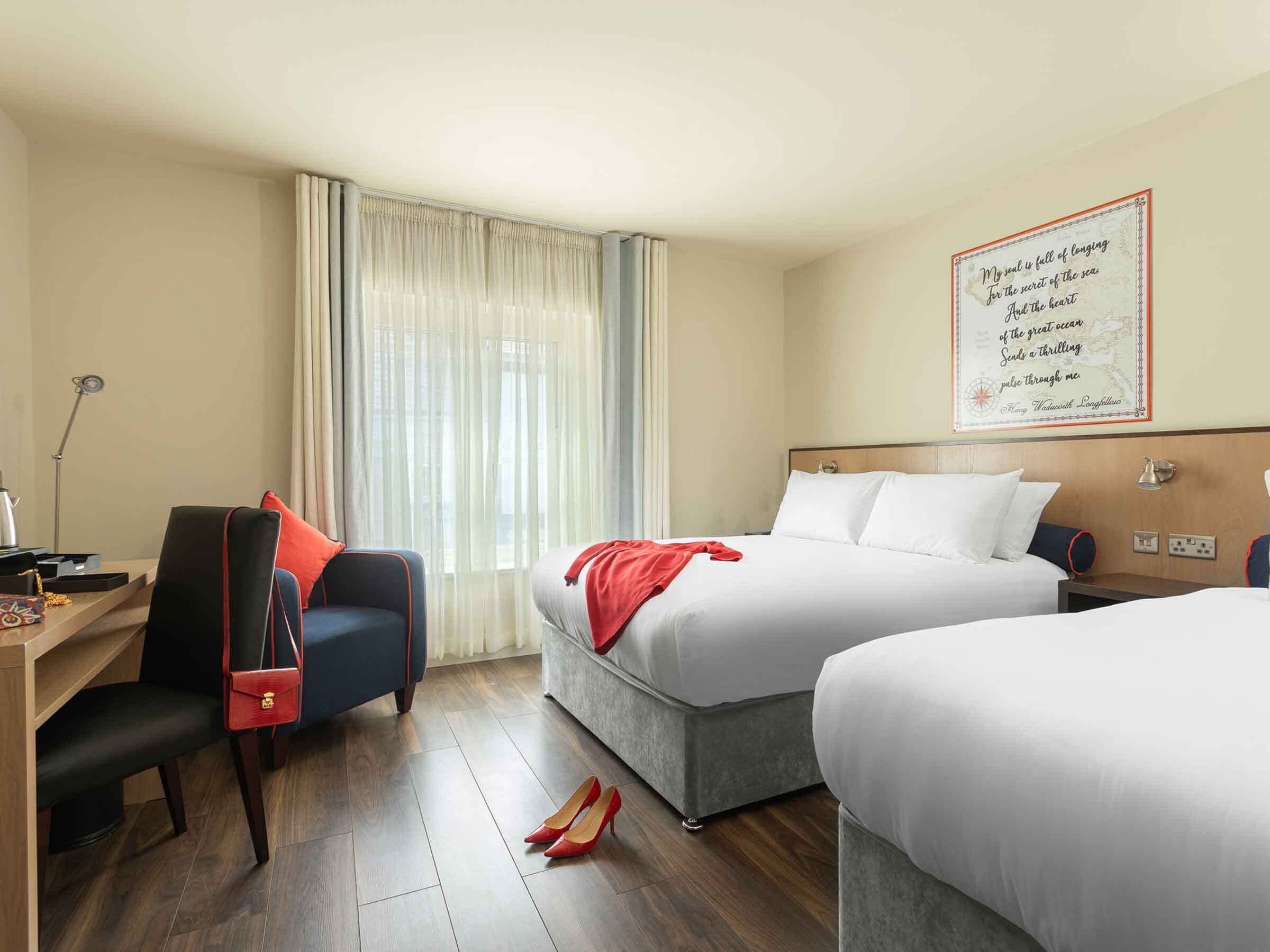Quarto de hotel com cama, escrivaninha, cadeira e janela com cortinas.