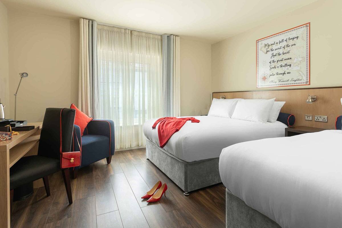 Quarto de hotel com cama, escrivaninha, cadeira e janela com cortinas.