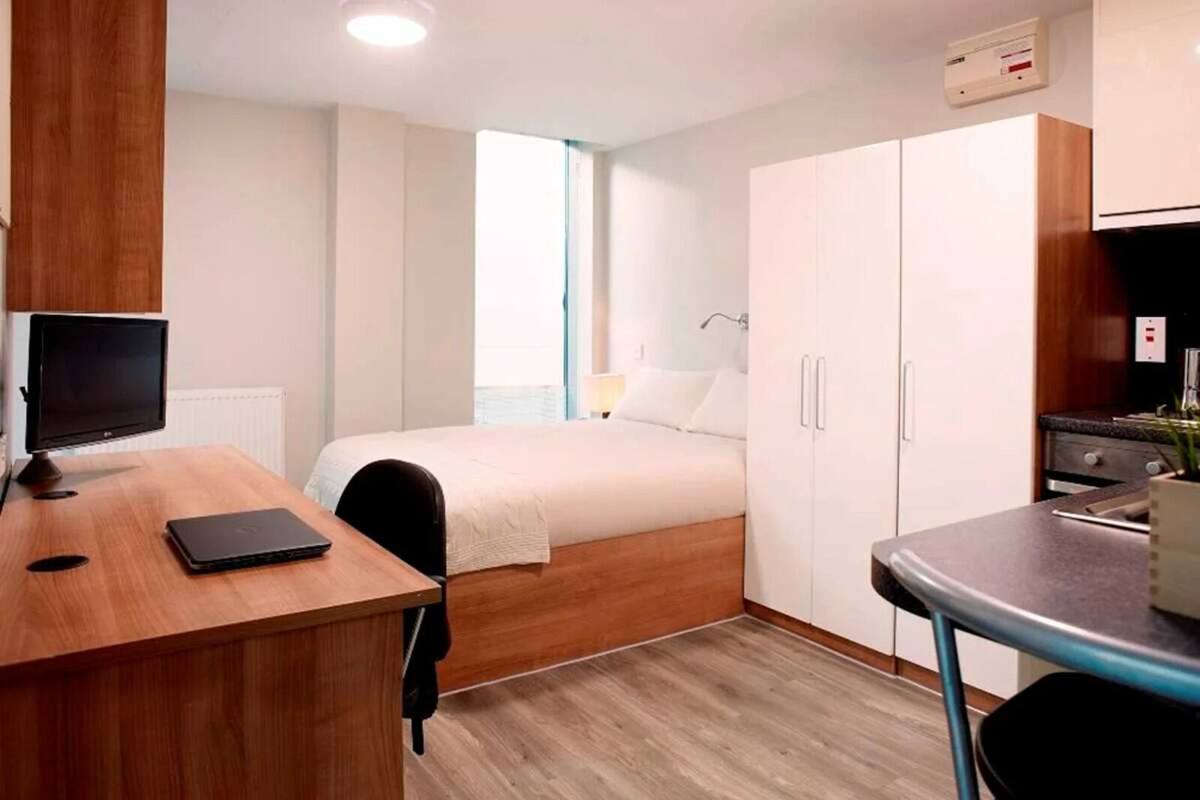 Quarto de hotel compacto com cama, escrivaninha com computador, televisão e cozinha compacta.