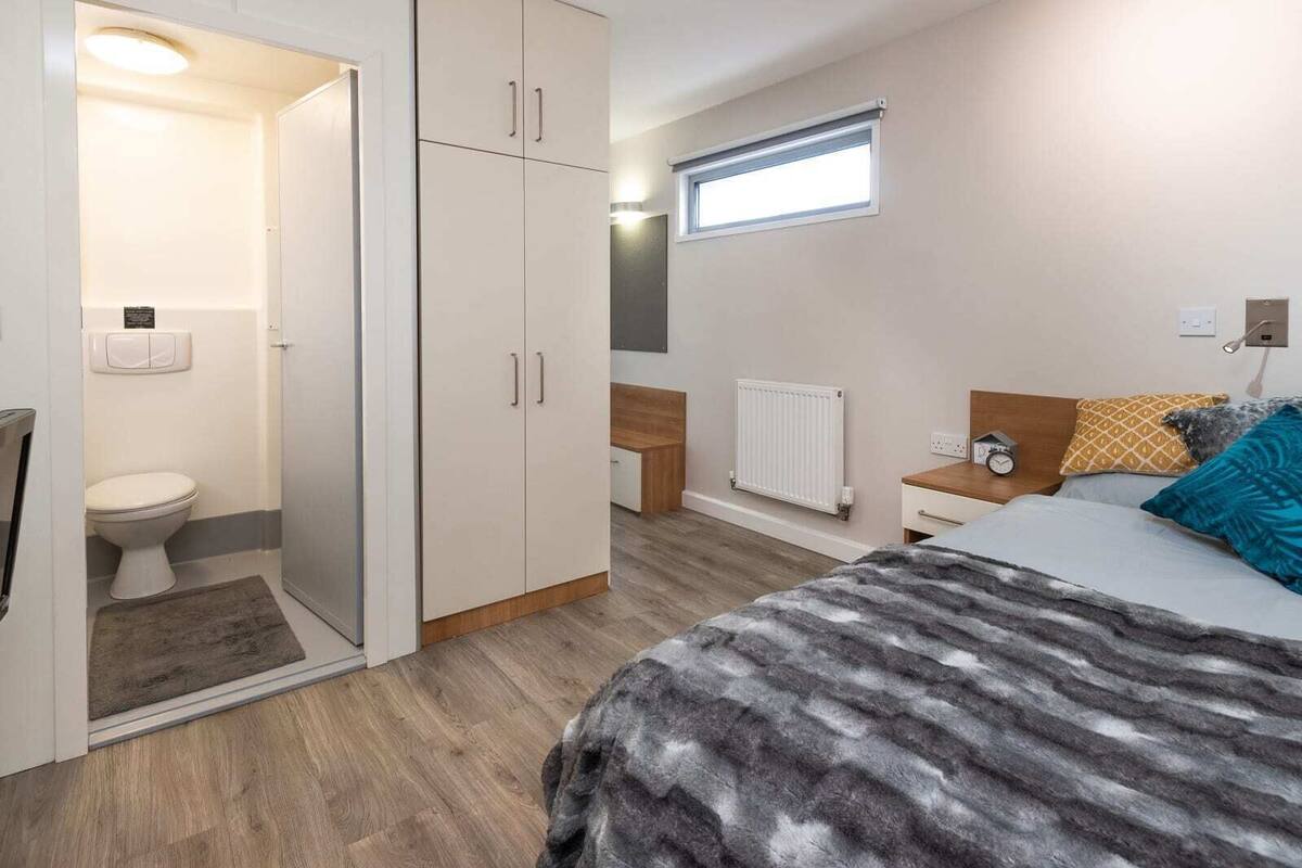 Um quarto com uma cama, mesa de cabeceira, janela, radiador e guarda-roupa.