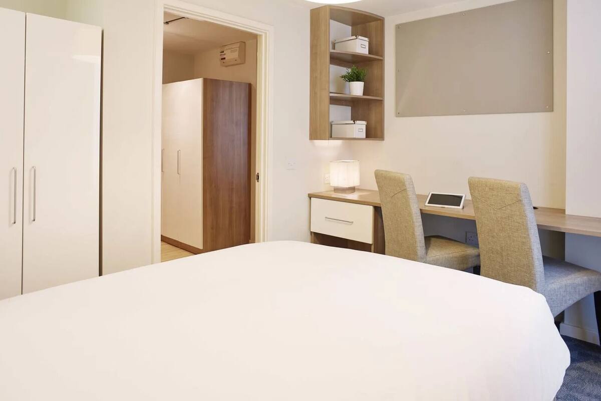 Quarto de hotel com uma cama, uma escrivaninha com cadeira, um guarda-roupa e uma prateleira com uma planta.