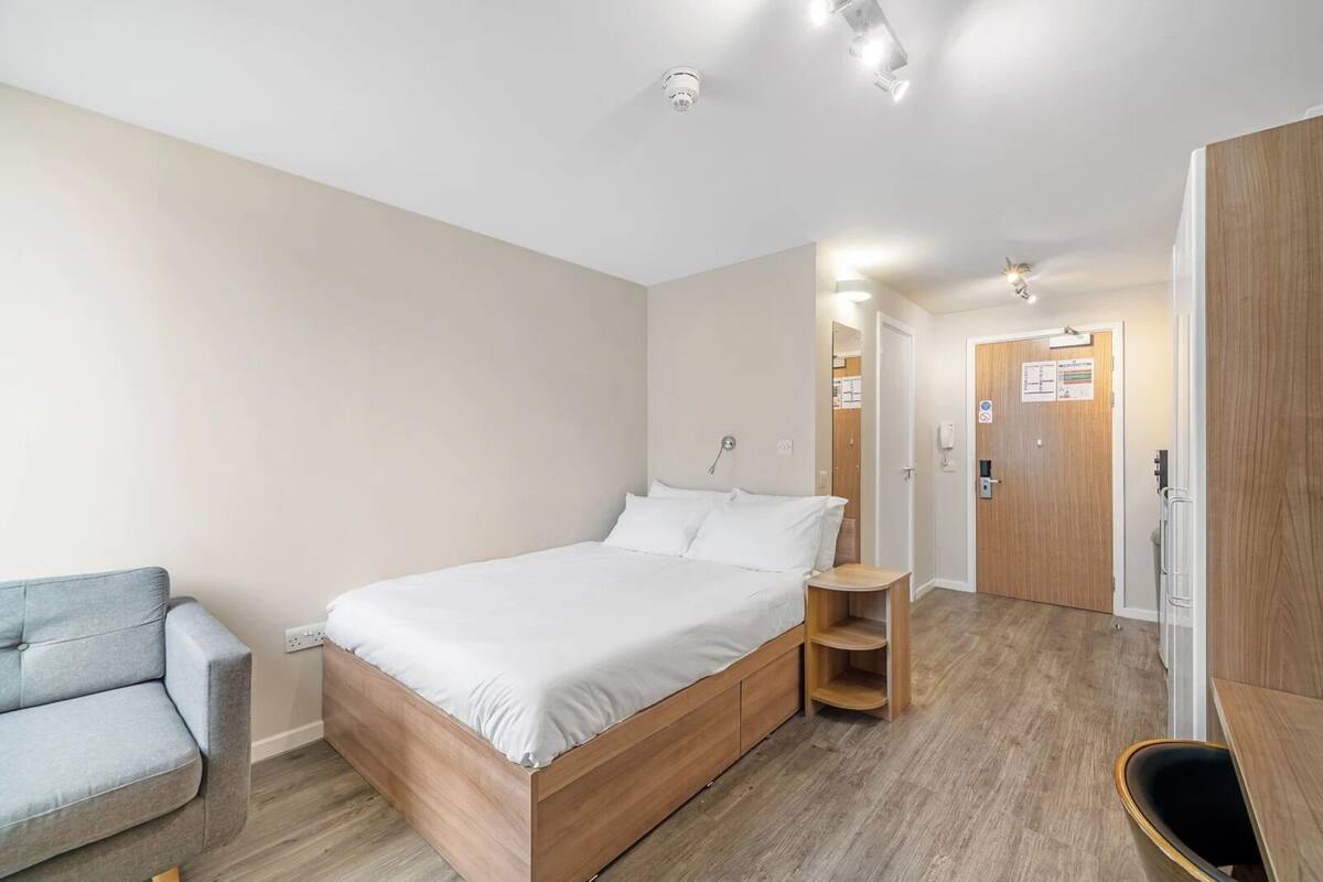 Quarto moderno com cama de madeira, poltrona cinza e pequena mesa de cabeceira de madeira.