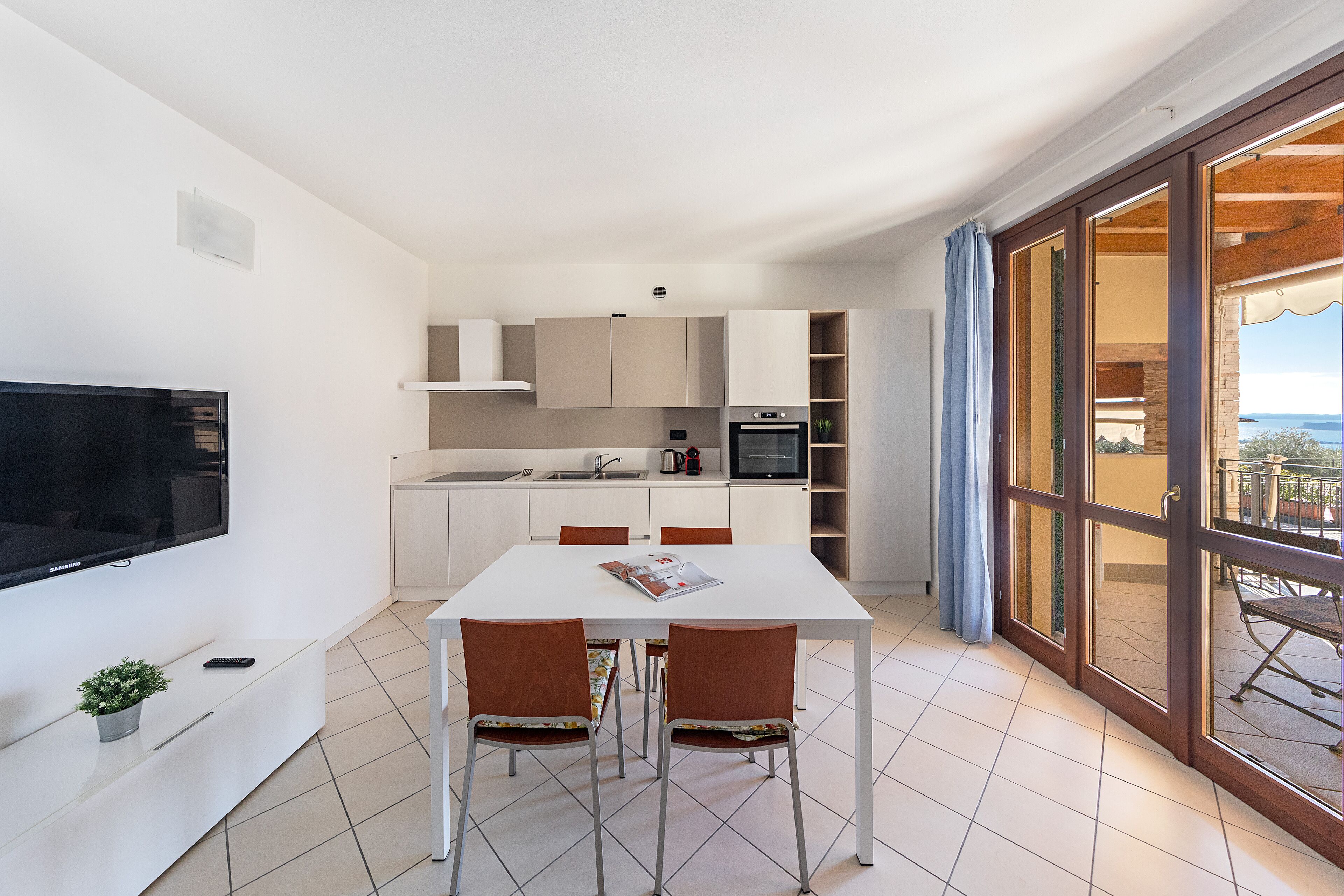 Cozinha moderna com armários brancos, mesa de jantar com quatro cadeiras, televisão e varanda com vista.