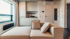 Quarto moderno com sofá, janela, cozinha compacta e porta.
