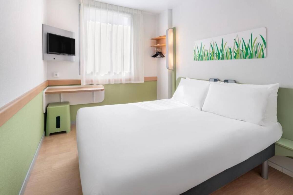 Quarto de hotel com cama, televisão na parede, um banquinho pequeno e uma pintura de grama.