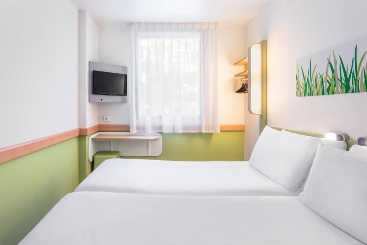 Quarto de hotel com cama, televisão, escrivaninha, cadeira, janela com cortinas e luminária na parede.
