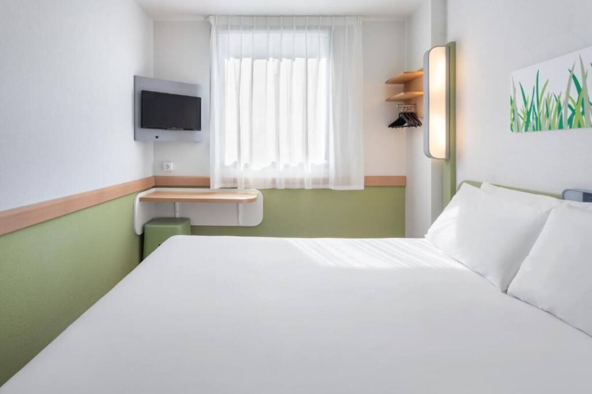 Quarto de hotel com cama, televisão fixada na parede, janela com cortinas translúcidas, pequena escrivaninha com cadeira e prateleira com sapatos.