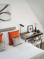 Quarto moderno com cama, almofadas, escrivaninha, luminária, um vaso com flores e um quadro emoldurado.