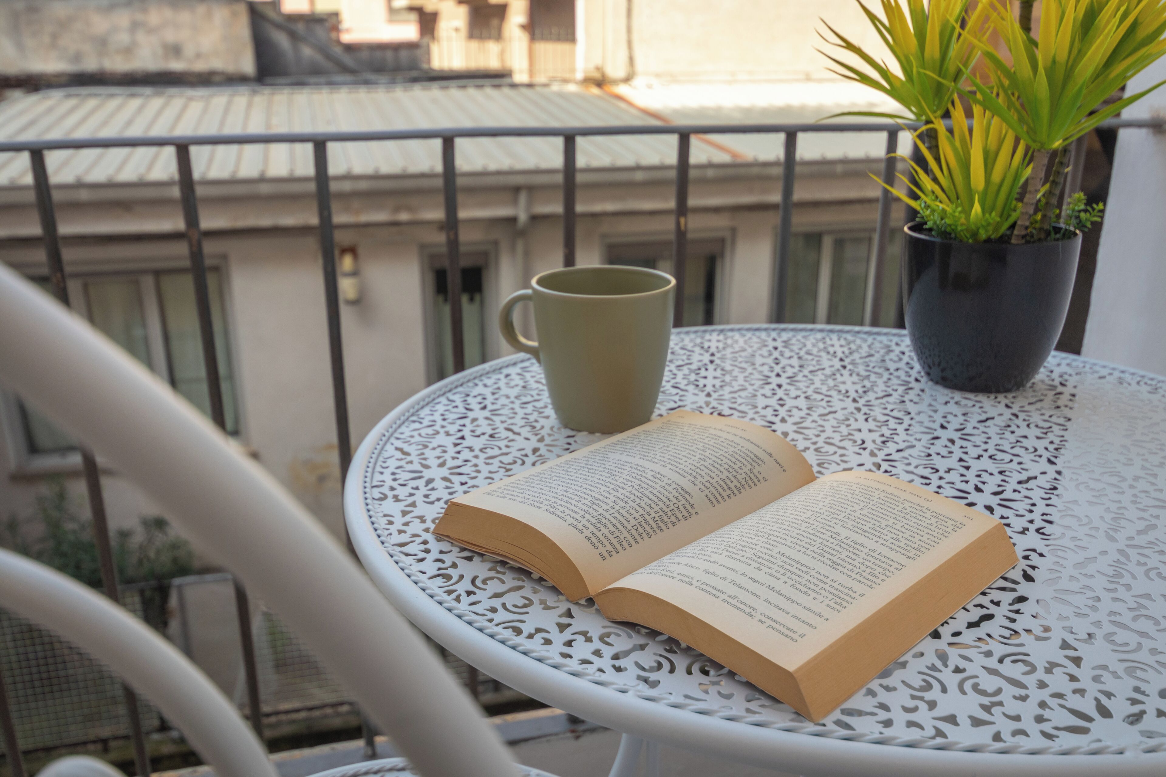 Um livro aberto sobre uma mesa de metal branca, com uma planta em vaso e uma caneca verde numa varanda.