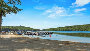 Um cais à beira do lago com barcos, uma praia de areia e colinas arborizadas ao fundo.