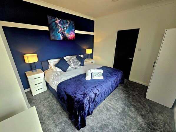 Quarto com cama, duas mesas de cabeceira, um criado-mudo, uma porta e uma obra de arte pendurada na parede.