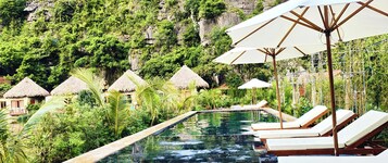 Uma área de piscina com espreguiçadeiras e guarda-sóis, rodeada por vegetação exuberante e palafitas com telhado de palha.