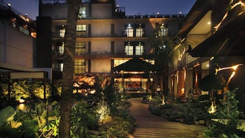 Pátio de hotel com caminho de madeira, palmeiras e um jardim de pedras.