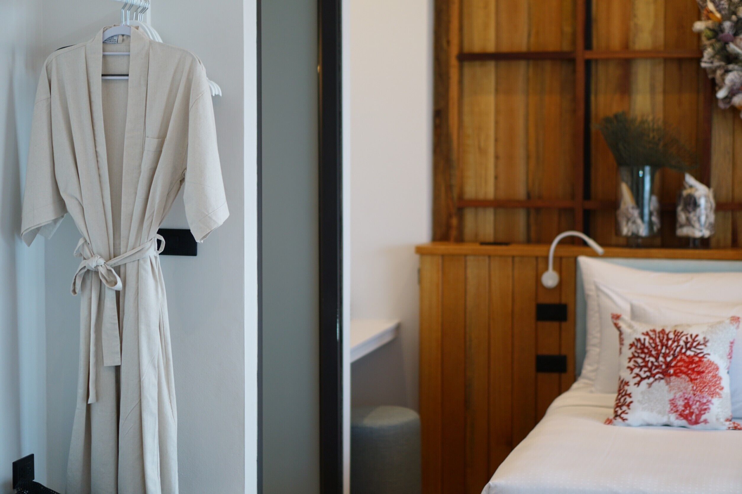 Quarto de hotel com armário de madeira, cama com roupa de cama branca e almofada com padrão coral, e um roupão pendurado em um suporte.