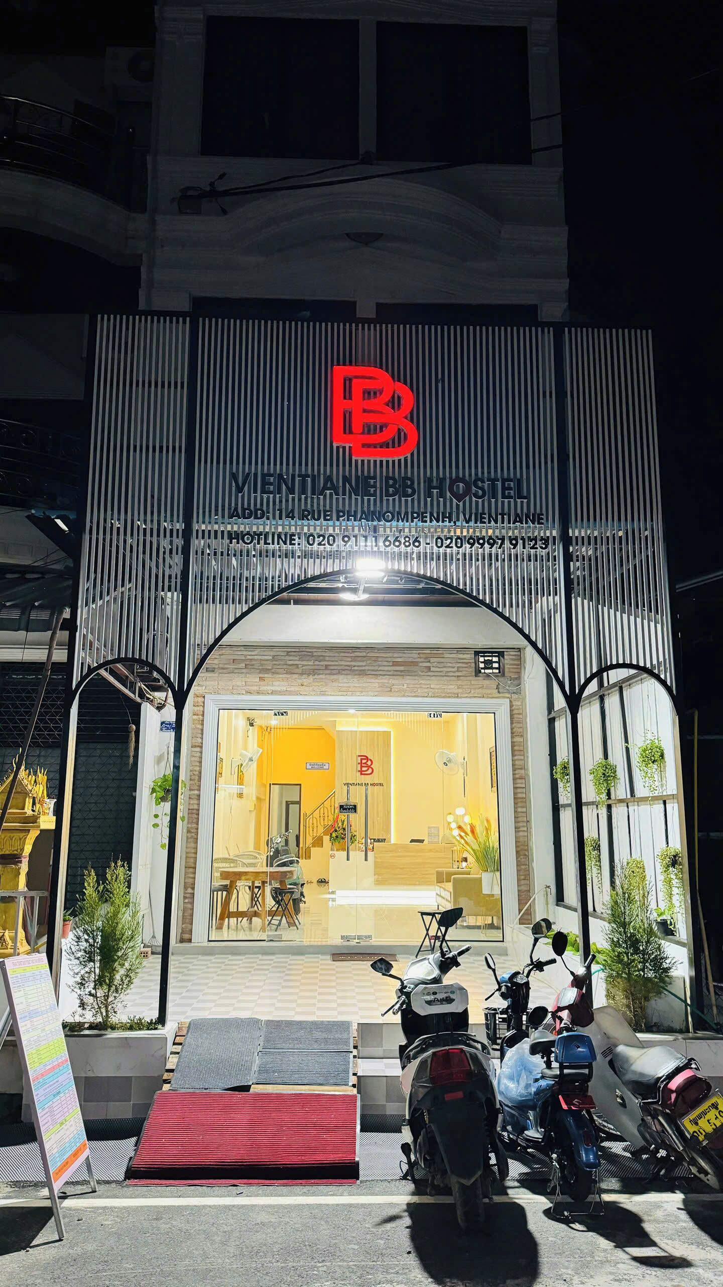 Um hostel com um logotipo vermelho chamativo, uma mesa de recepção e várias scooters estacionadas na frente.