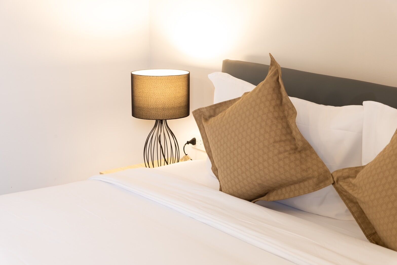 Uma cama bem arrumada com lençóis brancos e duas almofadas estampadas, uma luminária moderna com cúpula texturizada e uma cabeceira escura.