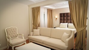 Quarto de hotel com um amplo sofá bege, uma poltrona combinando, uma cama com cabeceira estofada e cortinas bege.