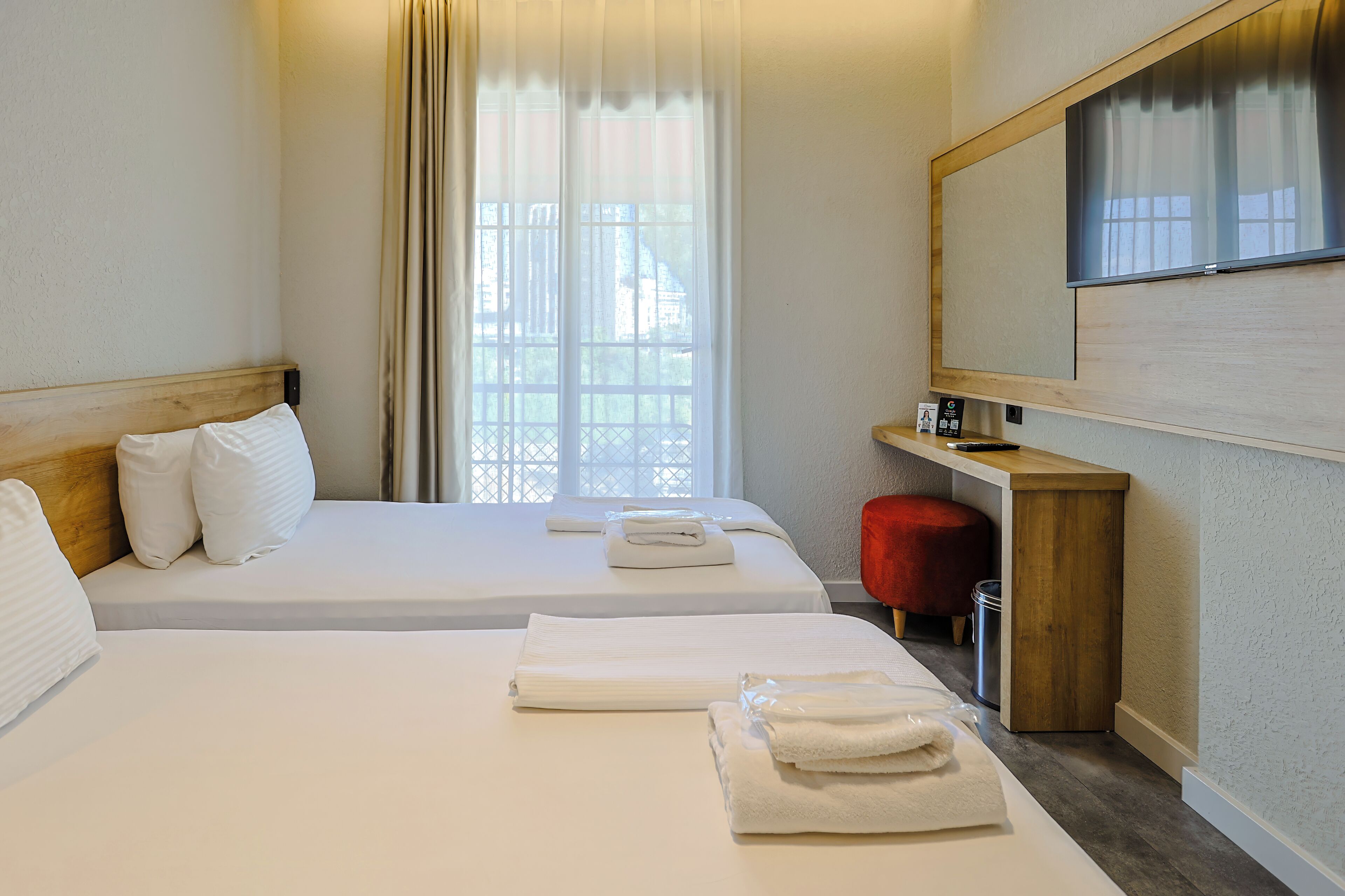 Quarto de hotel com duas camas, cabeceira de madeira, um banquinho vermelho, uma mesinha e uma televisão fixada na parede.