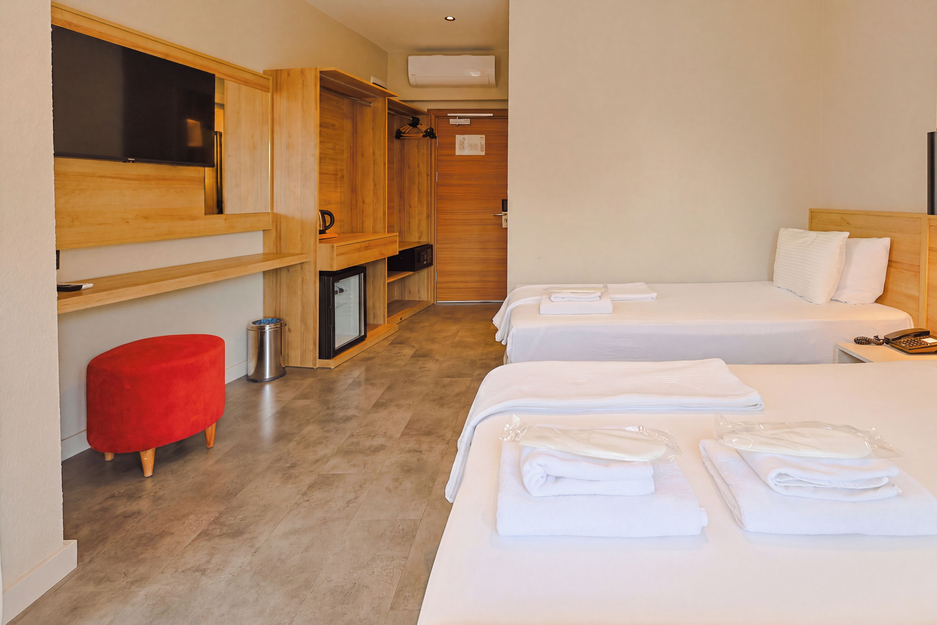 Quarto de hotel com duas camas, uma cadeira vermelha, uma kitchenette pequena e um guarda-roupa de madeira.