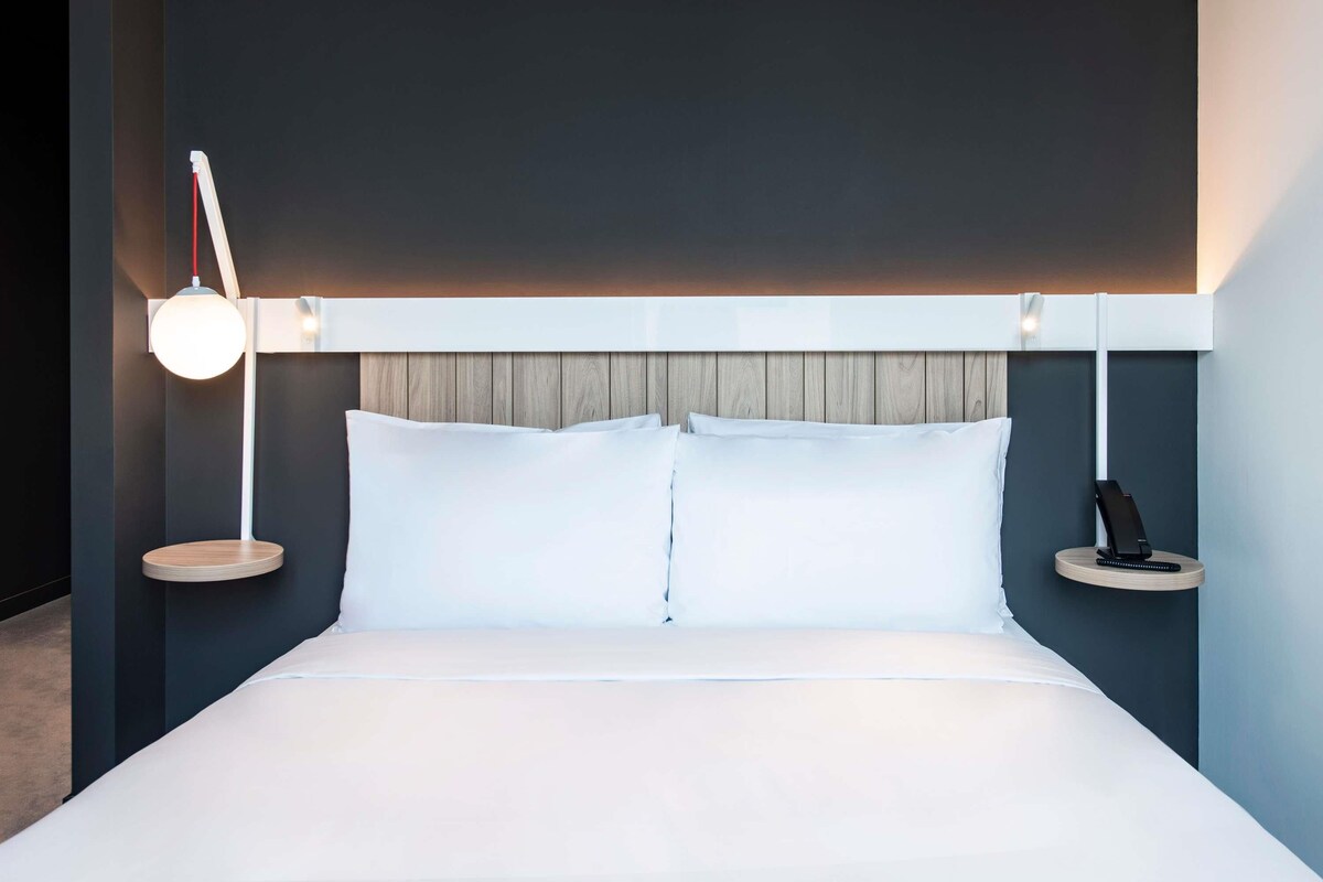 Quarto de hotel moderno com uma cama grande, abajur, telefone e uma mesinha.