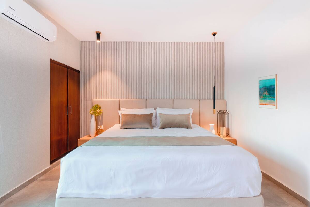 Quarto de hotel moderno com uma cama grande, um guarda-roupa de madeira, uma mesa de cabeceira com um vaso e um candeeiro fixado na parede.