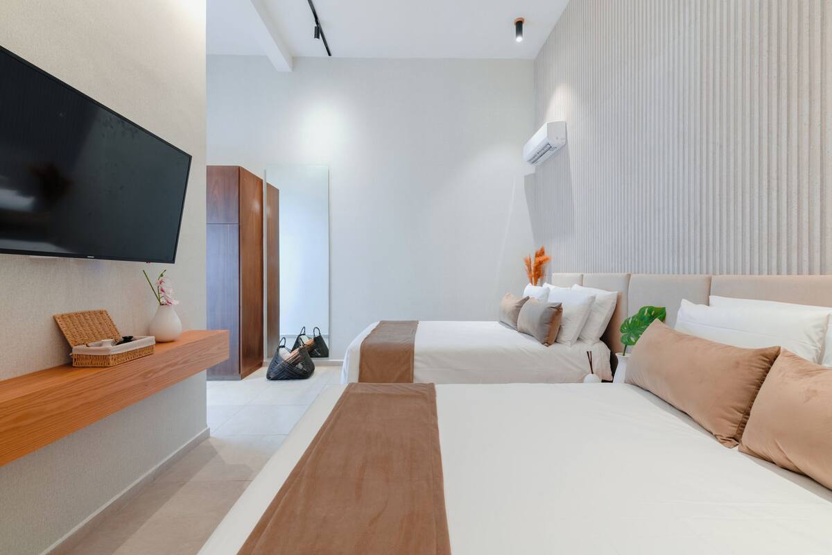 Quarto de hotel moderno com duas camas, uma televisão de tela plana na parede, uma prateleira de madeira com um cesto e um vaso, e uma janela grande com persianas verticais.