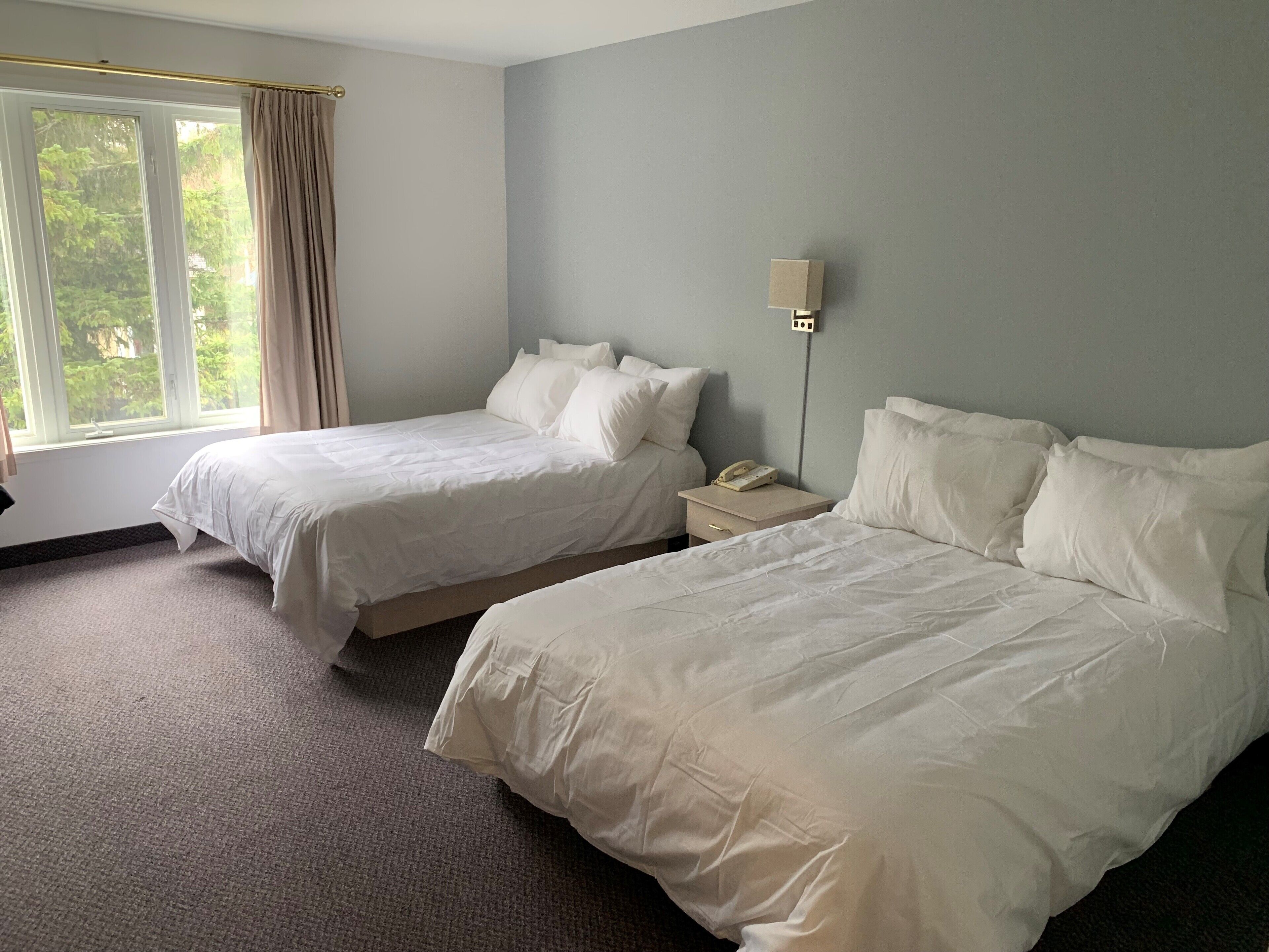 Quarto de hotel com duas camas, mesa de cabeceira com telefone e janela com cortinas.