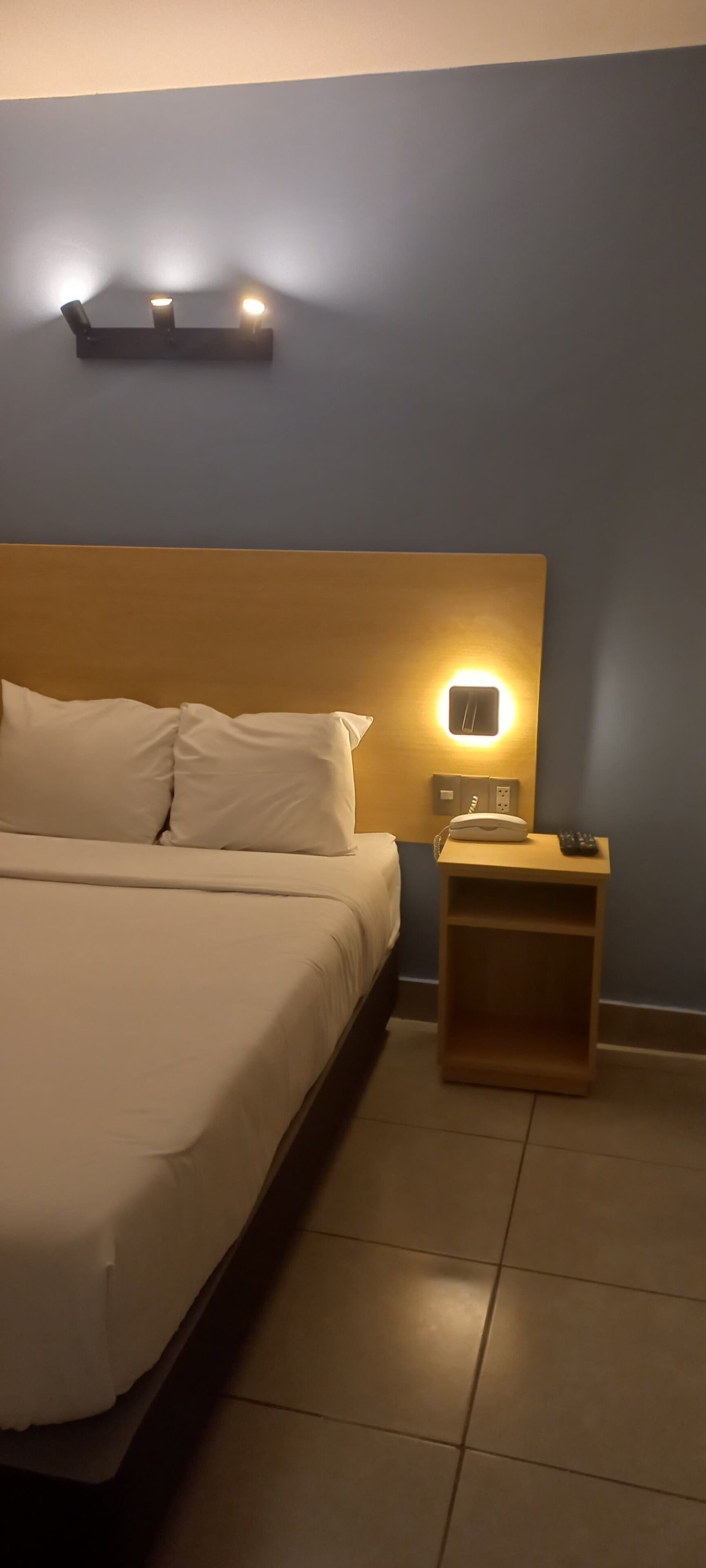 Quarto com cama, mesa de cabeceira com abajur e luminária na parede.
