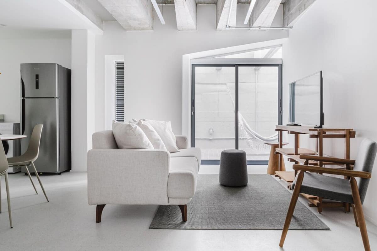 Sala de estar moderna com sofá, banquinho e cadeira de madeira. Uma janela grande oferece iluminação natural e uma porta deslizante dá acesso a uma varanda.