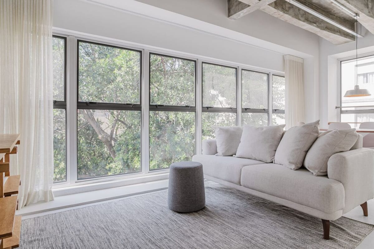 Uma sala de estar moderna com uma janela grande, um banquinho cinza e um sofá branco com várias almofadas.