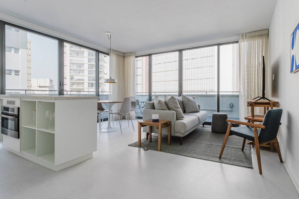 Sala de estar moderna com sofá, mesa de centro e cadeiras. Há uma área de cozinha com geladeira e uma pequena ilha. Grandes janelas oferecem vista da cidade.