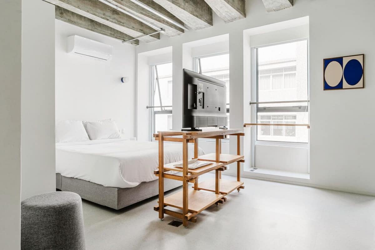Quarto moderno com cama, televisão sobre suporte de madeira e grandes janelas.