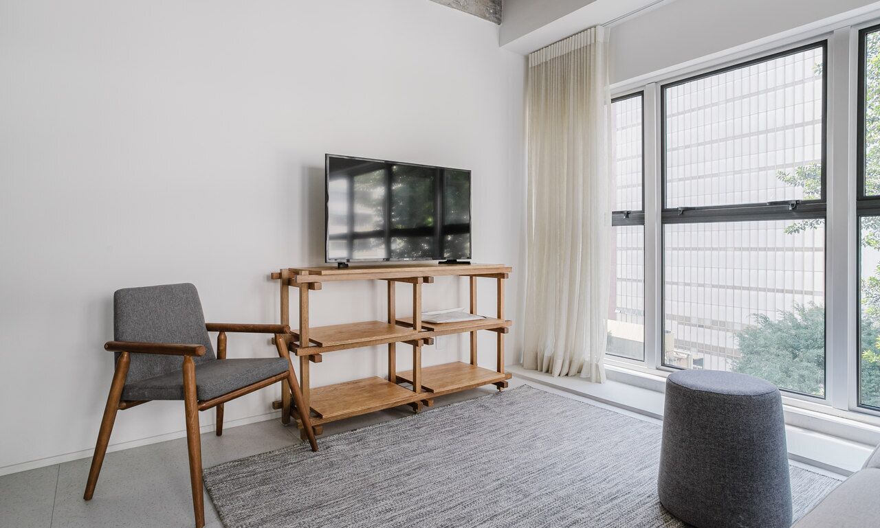Quarto moderno com televisão sobre suporte de madeira, uma poltrona cinza, um banquinho cinza e uma janela grande com vista para a cidade.