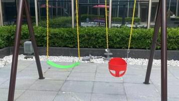 Um conjunto de baloiços com dois assentos, um verde e um vermelho, numa área pavimentada ao ar livre, com um viveiro e um edifício ao fundo.