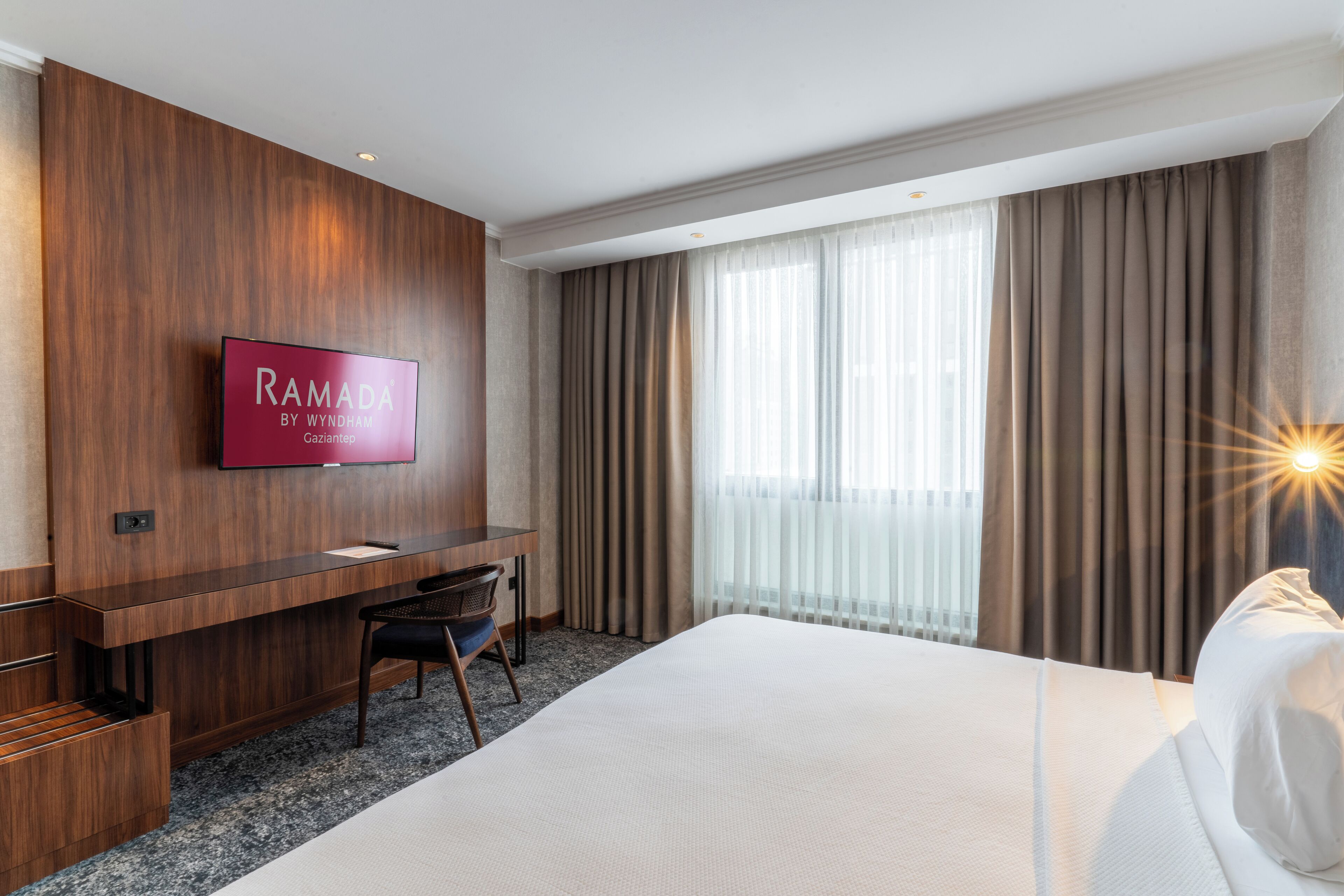 Quarto de hotel com cama, escrivaninha, cadeira, televisão presa na parede e uma janela grande com cortinas.