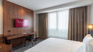 Quarto de hotel com cama, escrivaninha, cadeira, televisão presa na parede e uma janela grande com cortinas.