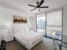 Quarto de hotel moderno com uma cama grande, duas mesas de cabeceira com luminárias, um ventilador de teto, vista para a cidade através de janelas do chão ao teto e um quadro na parede.