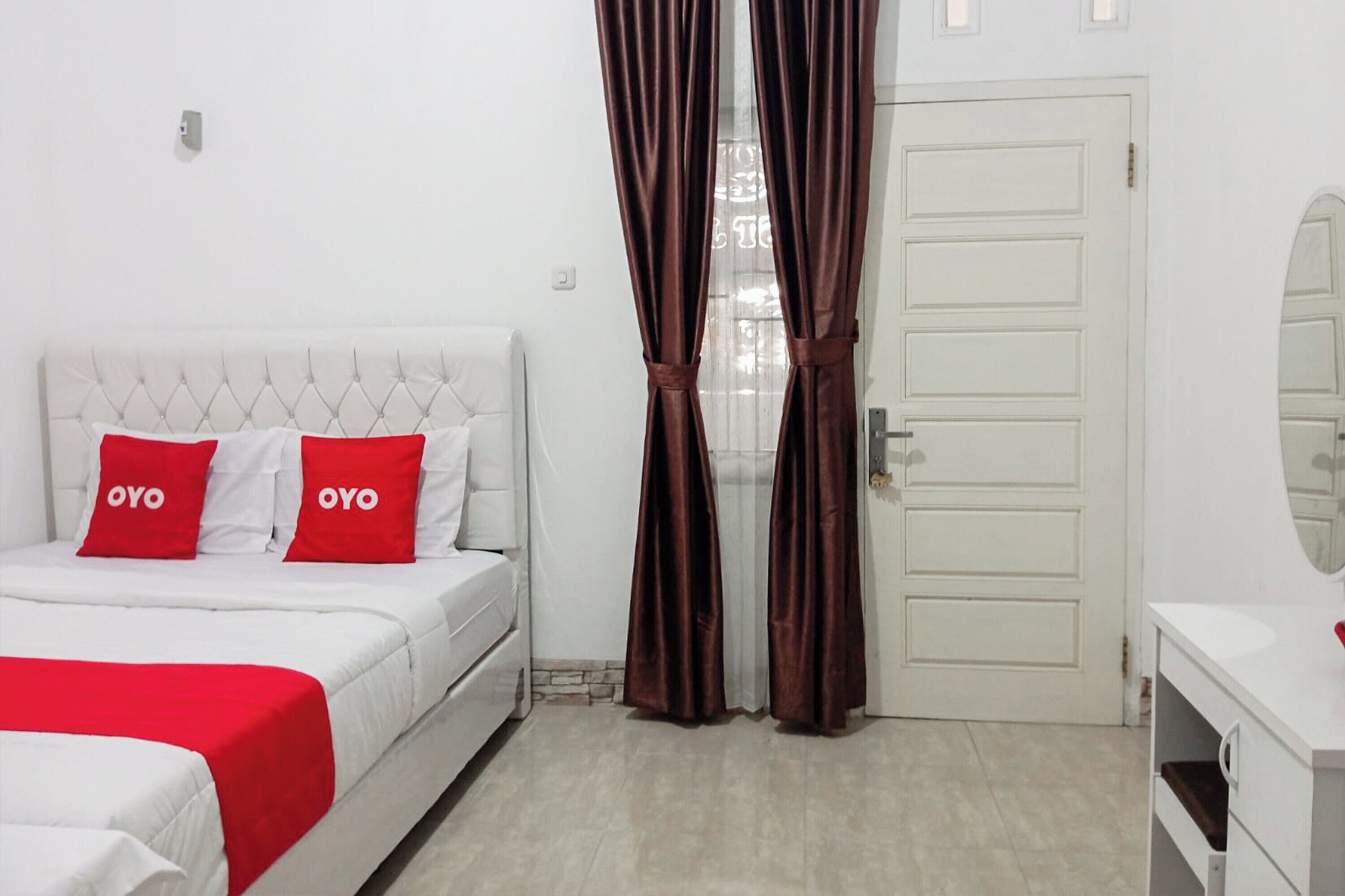 Quarto de hotel com cama branca, almofadas vermelhas, penteadeira branca e porta branca.