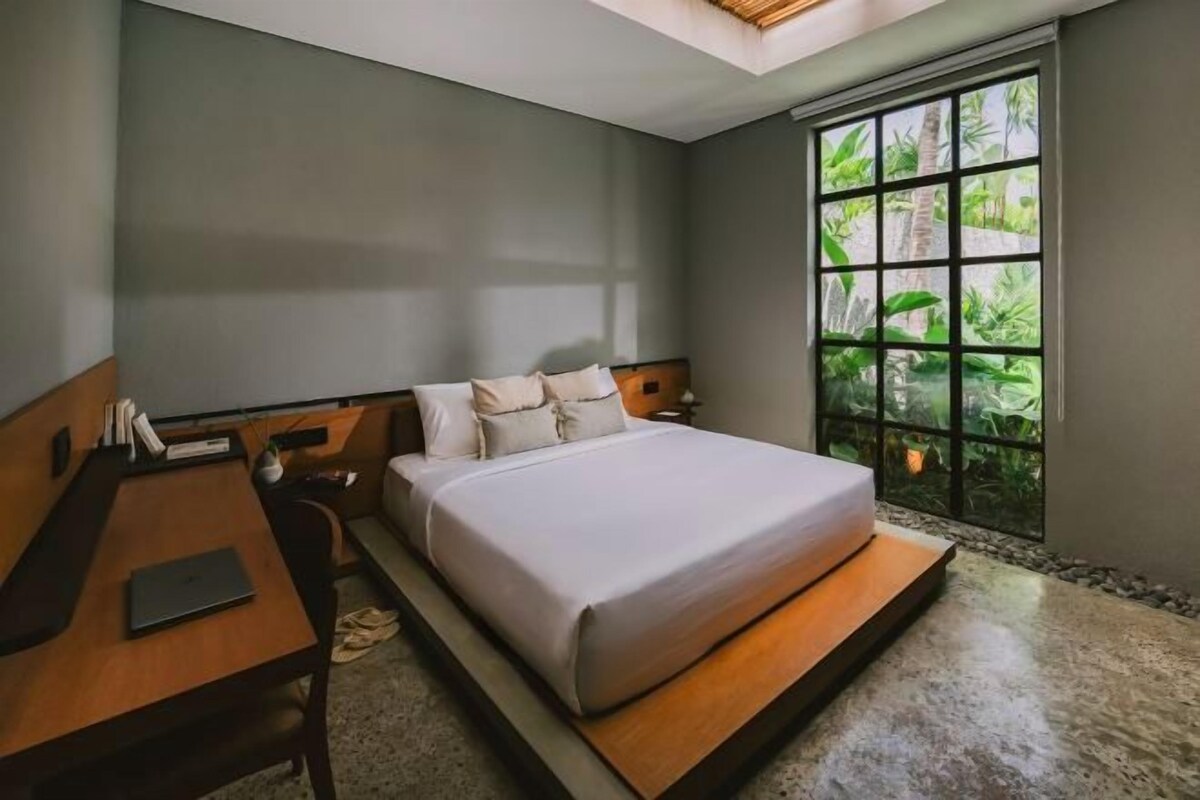 Quarto moderno com cama grande, escrivaninha e janela com vista para vegetação.