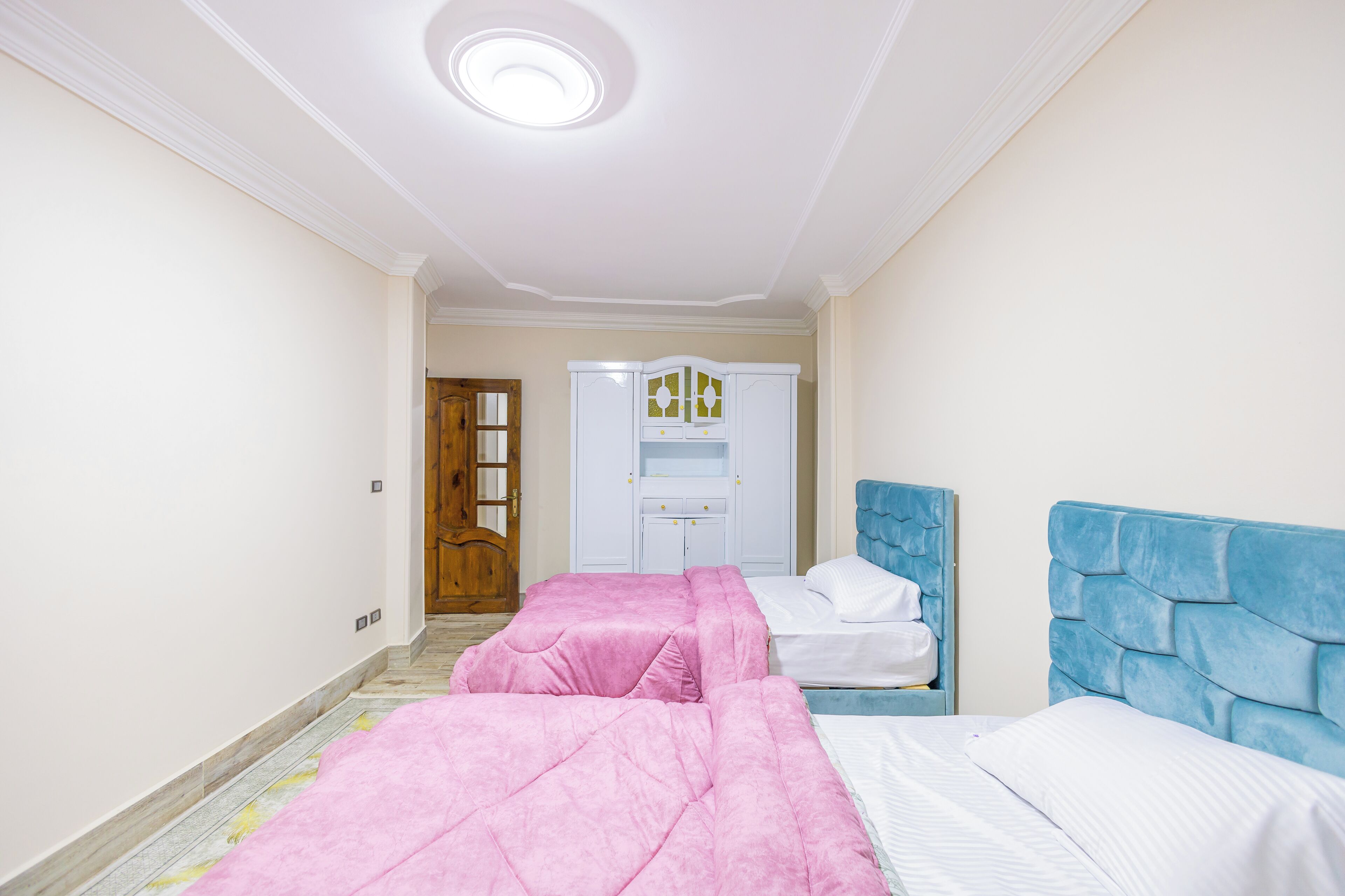Quarto de hotel com duas camas, uma com cabeceira azul e outra com colcha rosa.