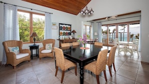 Uma sala de jantar ampla, com uma mesa grande e cadeiras de vime, teto de madeira e janelas grandes que oferecem vista para o exterior.