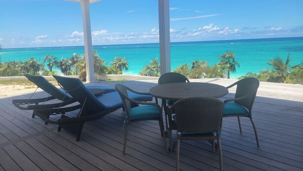 Uma área externa coberta com cadeiras de descanso e uma mesa, com vista para um oceano azul-turquesa e palmeiras.