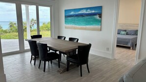 Uma área de refeições com mesa de madeira e cadeiras pretas, vista para a praia através de grandes janelas, e um quadro com uma cena de praia na parede.