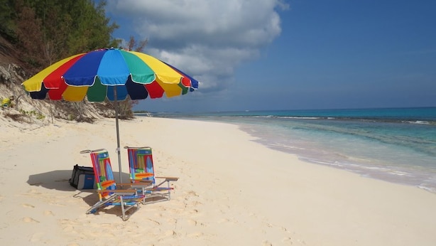 Uma praia com um guarda-sol colorido, duas cadeiras de praia e uma bolsa na areia.