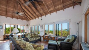 Uma sala de estar ampla com móveis de vime, grandes janelas com vista para o oceano e um ventilador de teto.