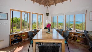 Uma sala de jantar ampla com uma longa mesa de madeira, cadeiras de vime e grandes janelas com vista para o oceano.