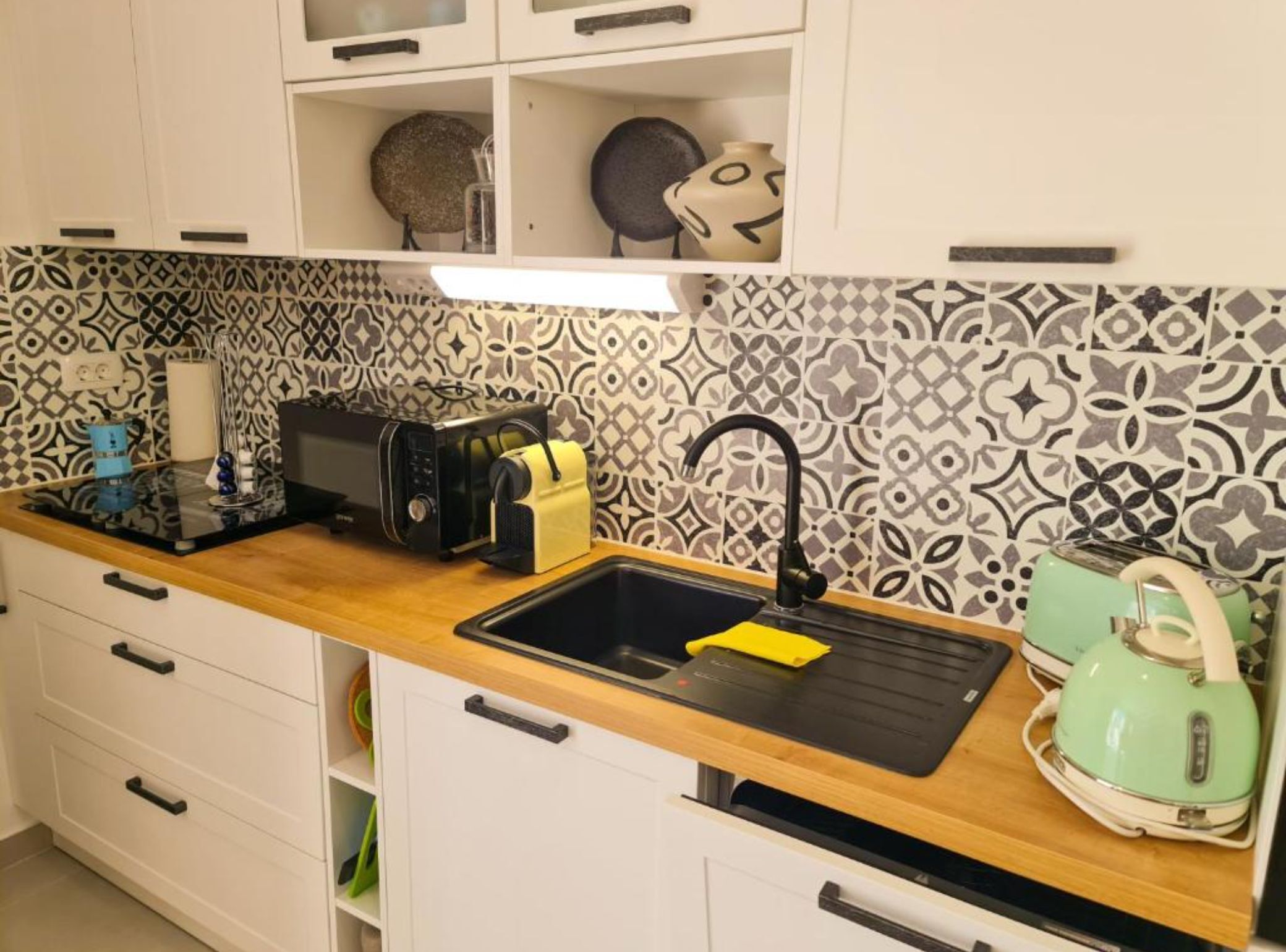 Uma cozinha com bancada de madeira, pia preta, chaleira verde, torradeira e painel de parede estampado.